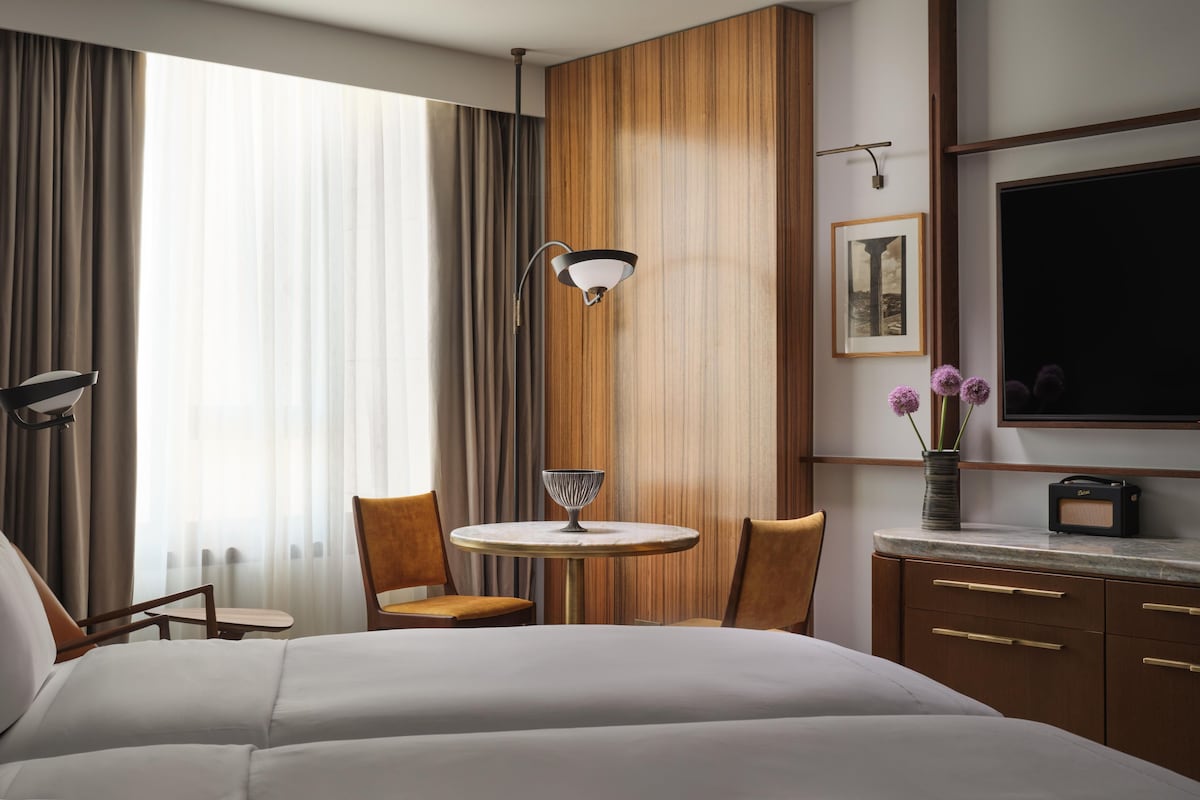 Quarto de hotel com uma cama grande, uma mesa redonda, duas cadeiras, uma escrivaninha com uma luminária, uma televisão e um quadro na parede.