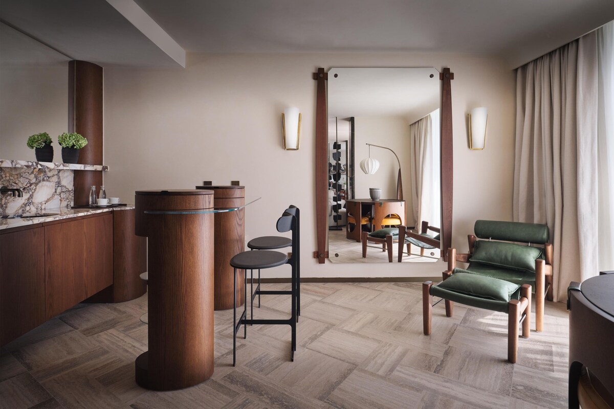 Quarto de hotel moderno com um balcão de madeira, área de estar com cadeiras, um espelho grande e padrão de piso em herringbone.