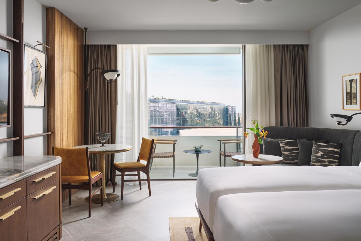 Quarto de hotel com uma cama grande, uma escrivaninha, uma cadeira, uma mesinha e vista para a cidade.