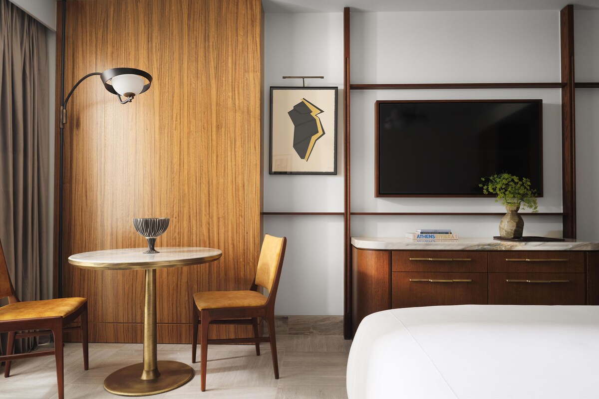 Quarto de hotel com parede de painel de madeira, uma mesa redonda com uma tigela, duas cadeiras, uma TV, um vaso com uma planta e uma cama.