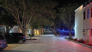 Vista noturna de um caminho de paralelepípedos que leva a um edifício com fachada branca e um carro preto estacionado à esquerda.