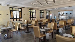 Um restaurante com várias mesas e cadeiras, grandes janelas e uma área de cozinha visível ao fundo.