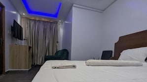 Quarto de hotel com uma cama grande, uma televisão de tela plana fixada na parede, uma cabeceira de madeira e iluminação LED azul no teto.