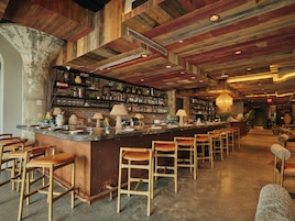 Uma área de bar com banquetas de madeira, um balcão e prateleiras repletas de garrafas.