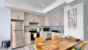 Cozinha moderna com armários de madeira clara, eletrodomésticos de aço inoxidável, uma mesa de jantar de madeira e um objeto decorativo na parede.