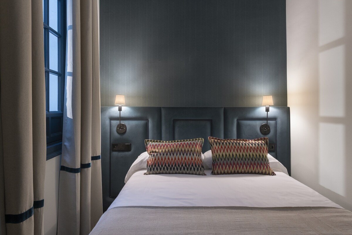 Quarto de hotel com cama, duas luminárias fixadas na parede e uma janela com cortinas.