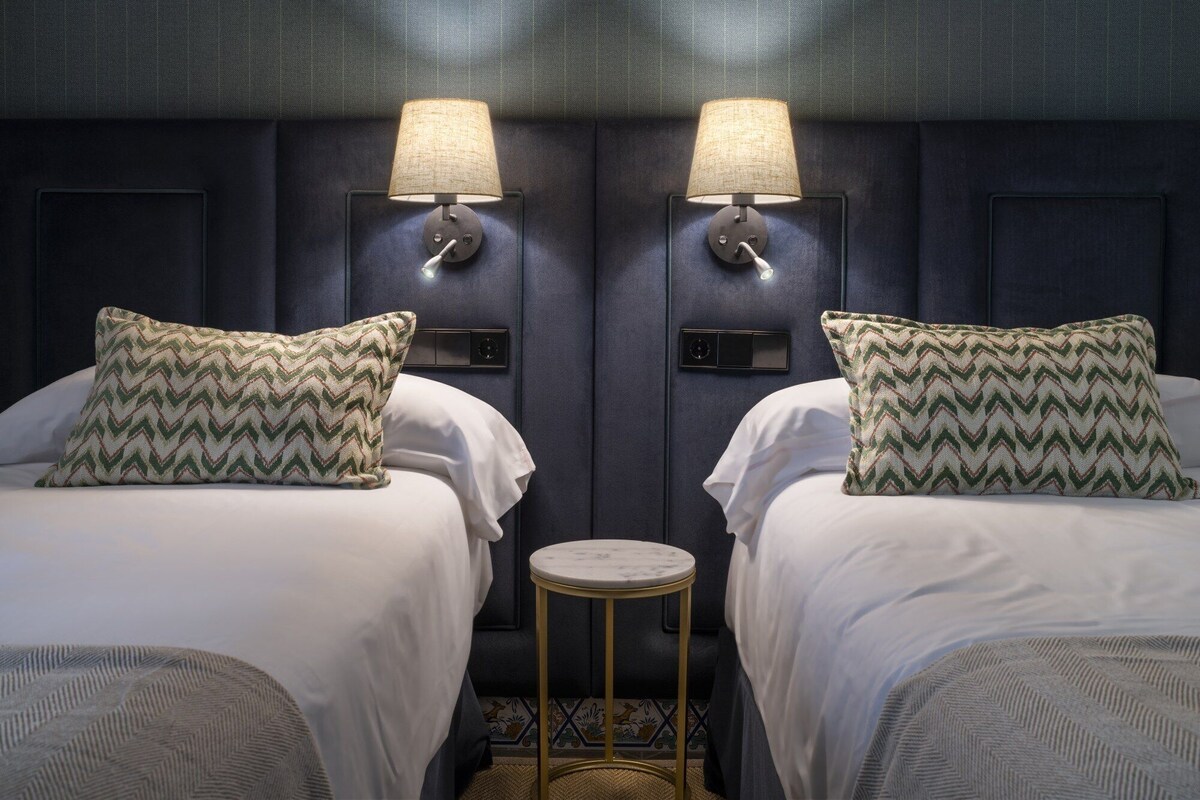 Quarto de hotel com duas camas, cada uma com almofada estampada, uma mesa redonda pequena e luminárias fixadas na parede.