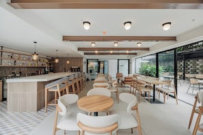 Um café moderno com área de bar, mesas e cadeiras de madeira e grandes janelas com vista para o verde.