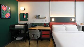 Quarto de hotel com cama, escrivaninha, cadeira e uma prateleira na parede com livros.