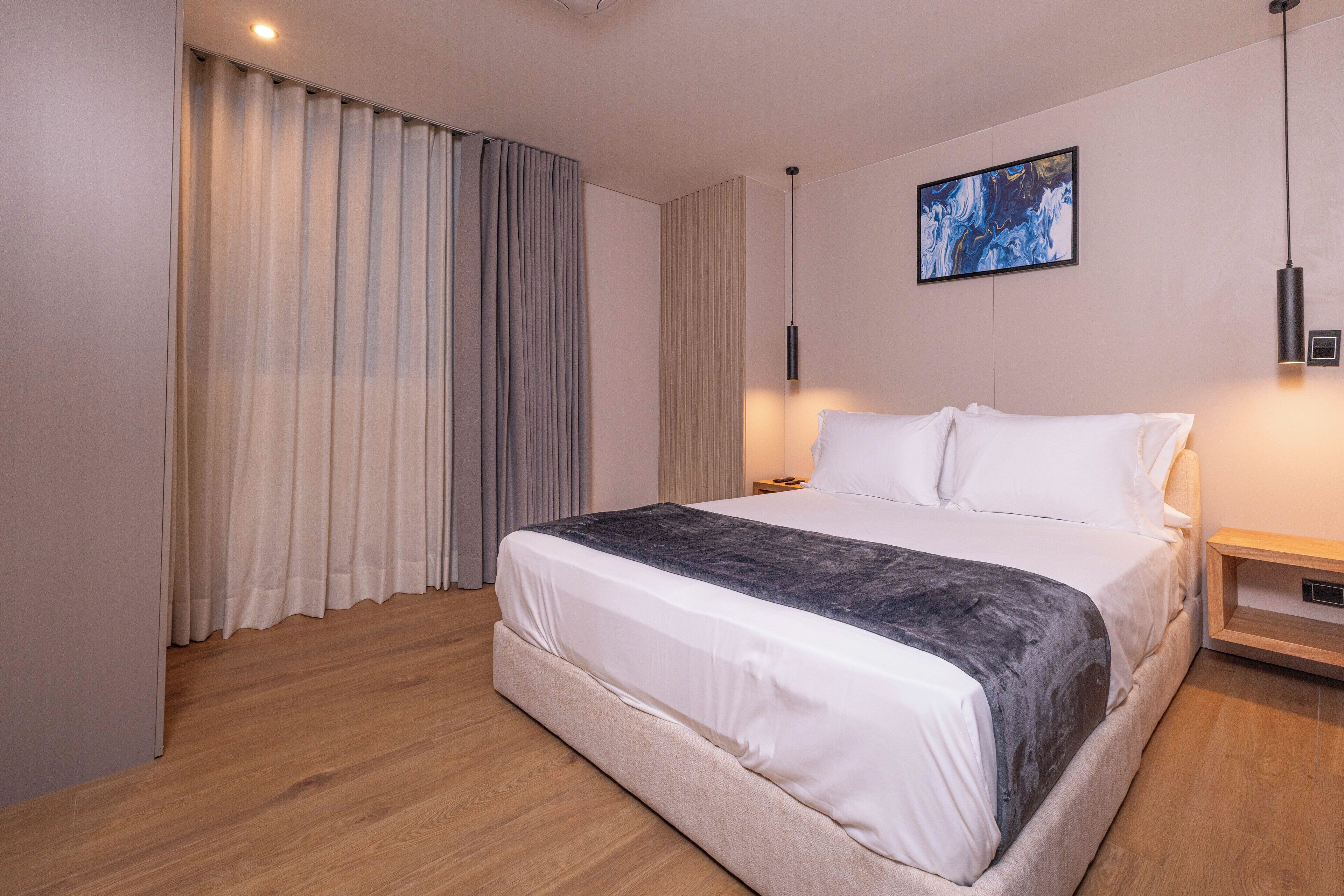 Quarto de hotel moderno com uma cama grande, piso de madeira, mesa de cabeceira, luminária na parede e um quadro.