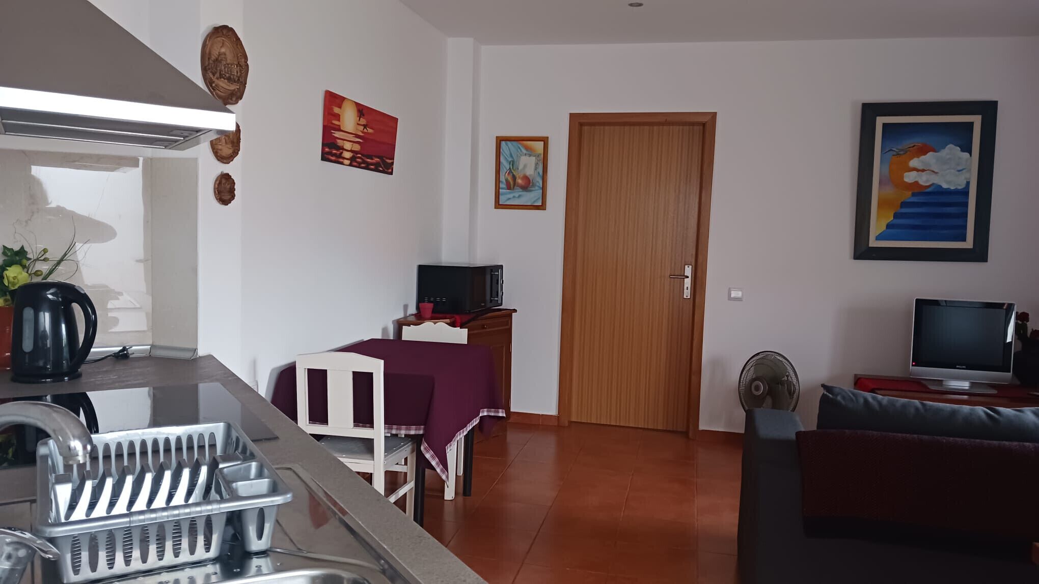 Cozinha moderna com pia, micro-ondas e área de jantar com mesa e cadeiras. Há uma porta de madeira, uma TV e um ventilador na sala.