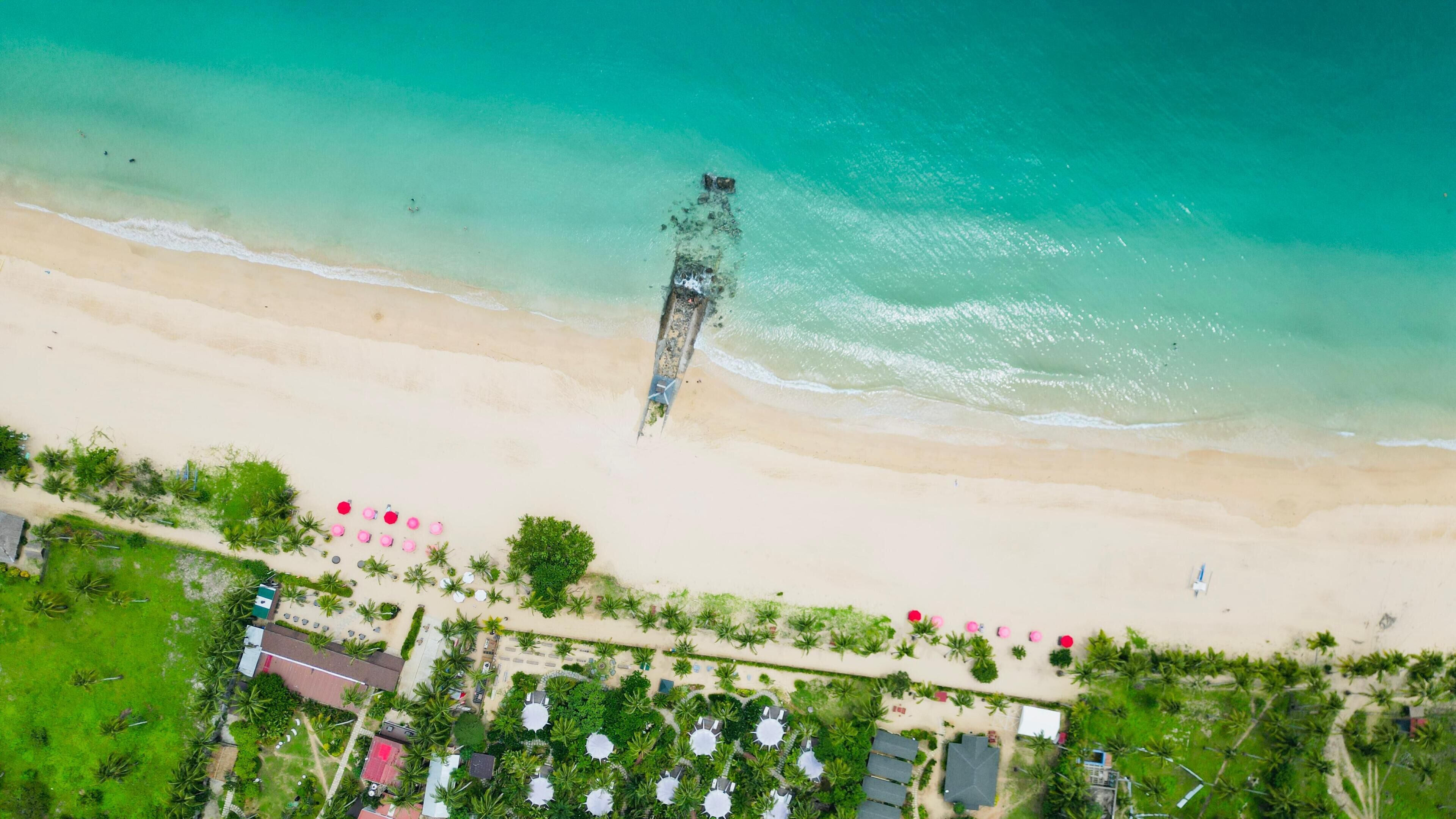 Vista aérea de uma praia com um píer que se estende na água, cercada por uma área de resort com vários edifícios e uma fileira de guarda-sóis de praia.