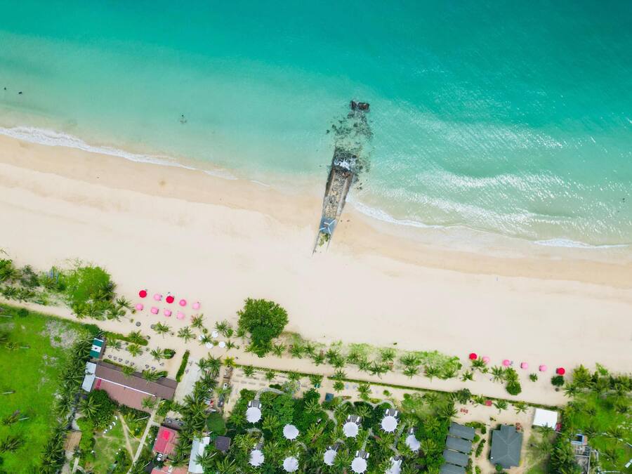Vista aérea de uma praia com um píer que se estende na água, cercada por uma área de resort com vários edifícios e uma fileira de guarda-sóis de praia.