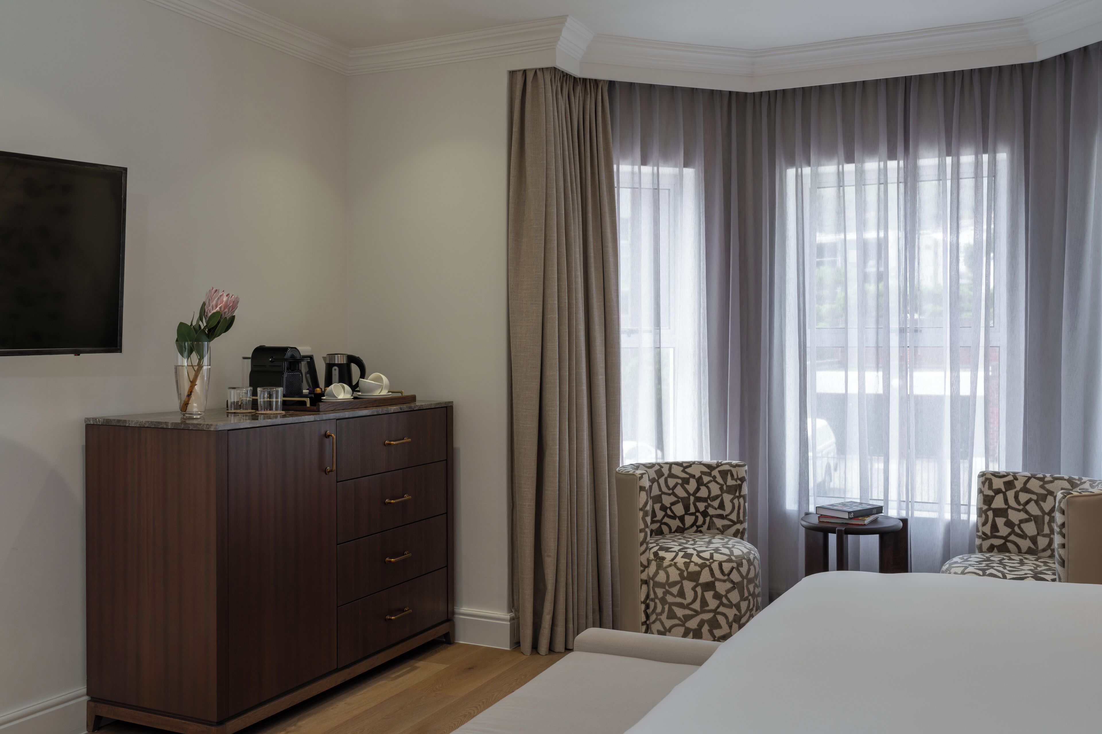 Quarto de hotel com uma cama grande, penteadeira de madeira, duas cadeiras estampadas, uma mesinha com livros e uma janela ampla com cortinas.
