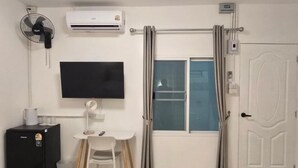 Quarto com TV de tela plana, uma pequena escrivaninha com cadeira, um mini-frigorífico preto, ar condicionado, janela com cortinas e uma porta branca.