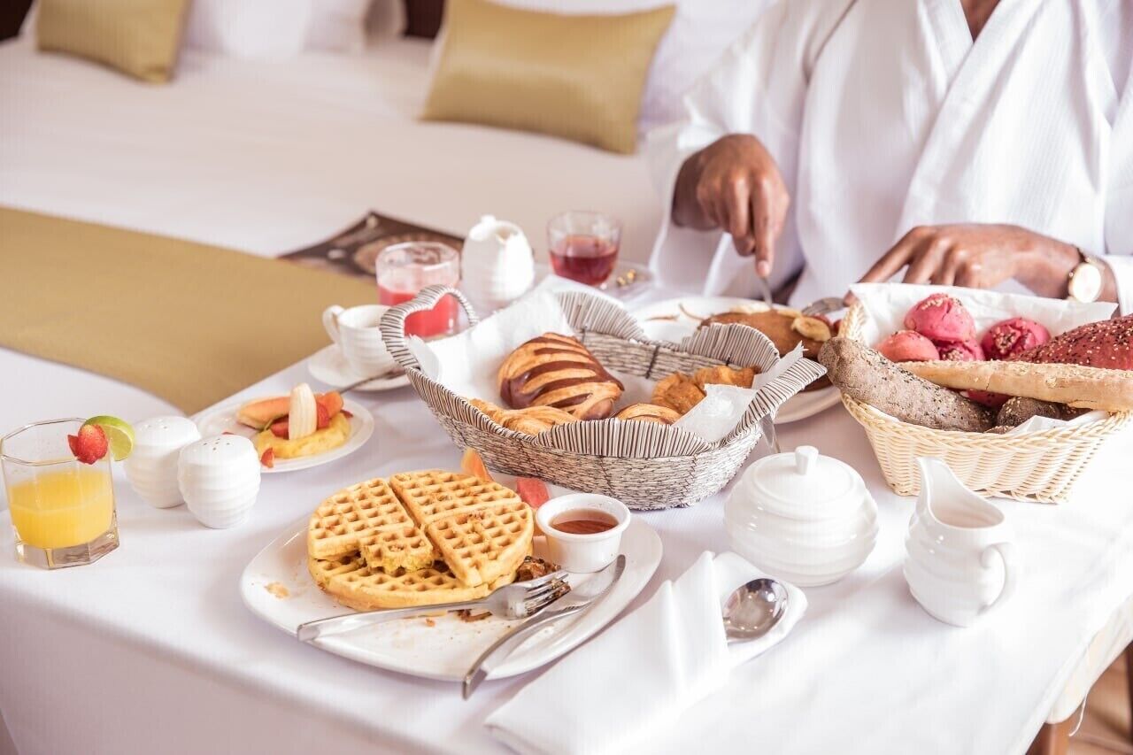 Mesa de café da manhã com waffles, doces e frutas, com uma cama ao fundo.