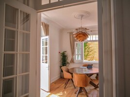 Porta aberta que dá para uma sala de jantar com uma mesa redonda, cadeiras e um lustre.