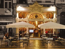 Um edifício ornamentado com um letreiro luminoso que diz “MAJESTIC” e o número “112” acima da entrada. Há mesas e cadeiras ao ar livre sob guarda-sóis brancos, e pessoas sentadas e circulando.