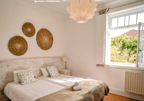 Quarto com cama grande, mesa de cabeceira, luminária na parede e vista para o verde lá fora.