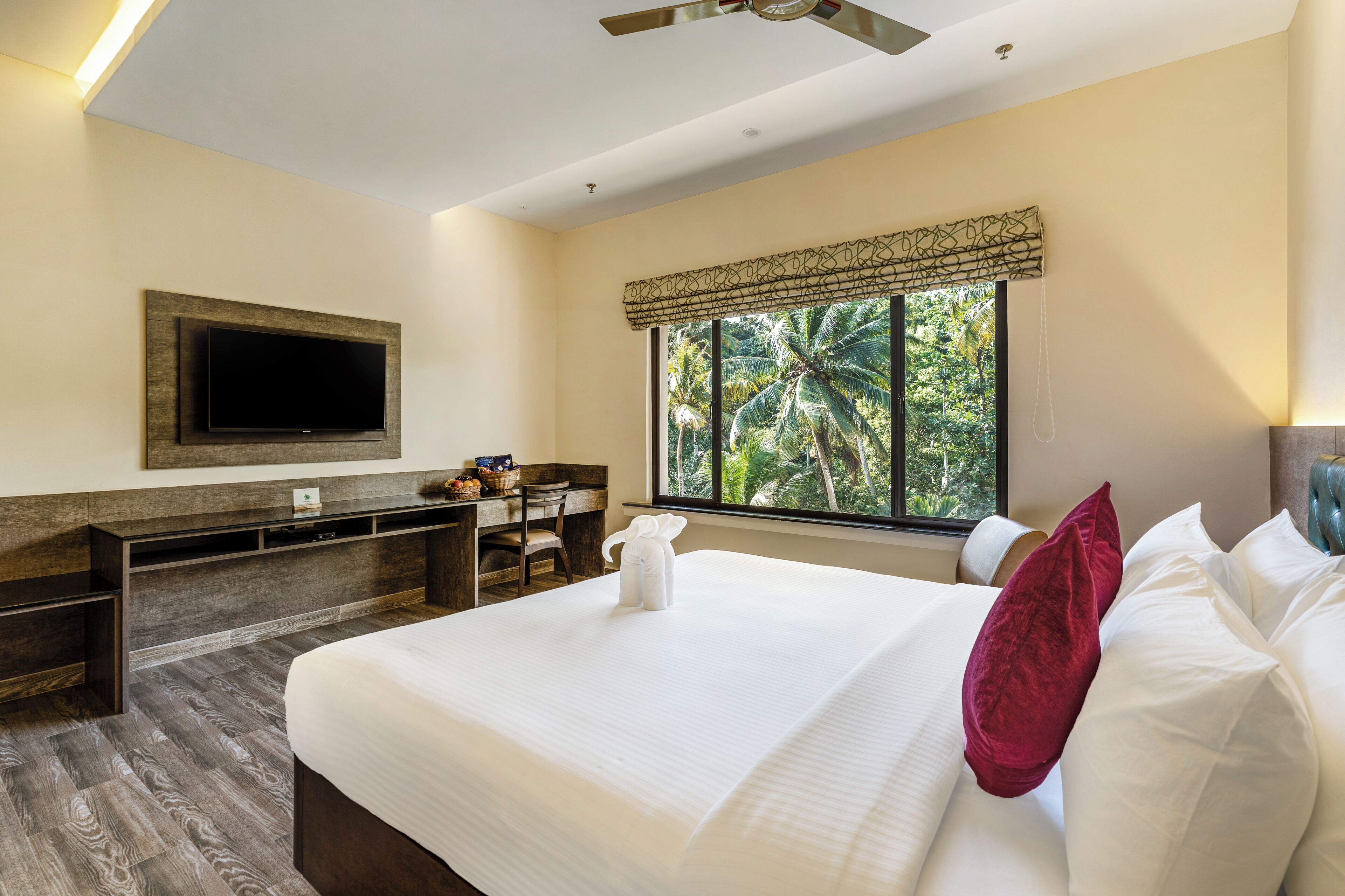 Quarto de hotel com uma cama grande, uma mesa de trabalho com televisão de tela plana, uma janela com vista para palmeiras e um ventilador de teto.