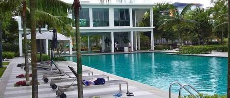 Piscina com cadeiras de descanso e equipamentos de ginástica, cercada por palmeiras e um edifício moderno.