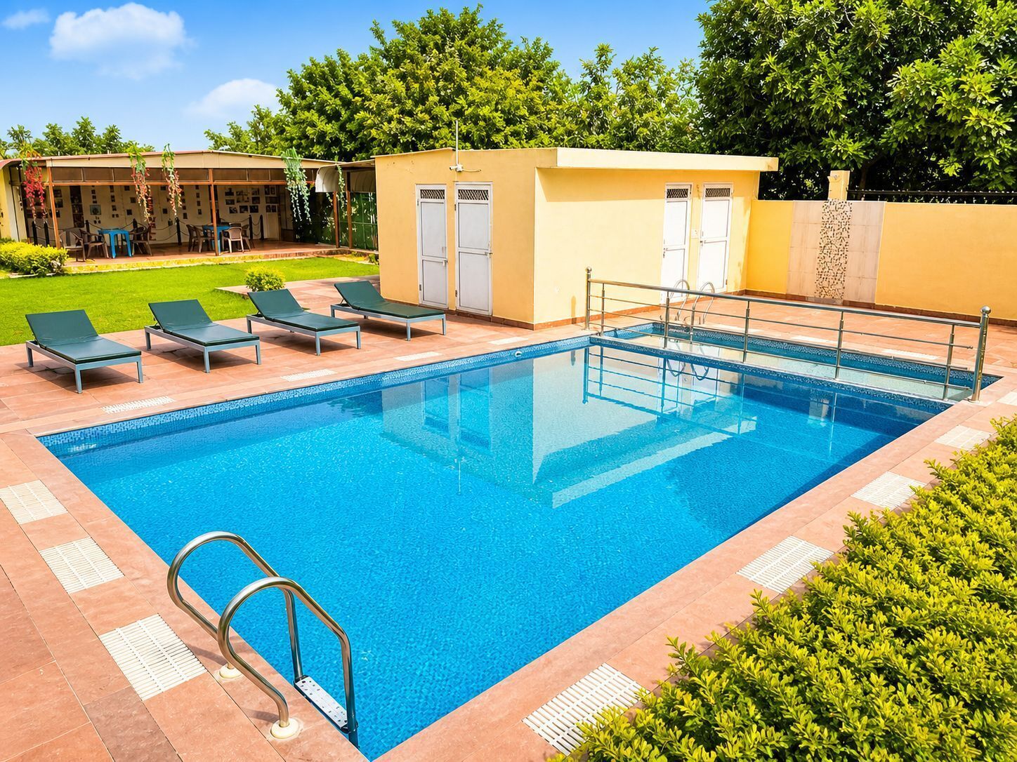 Piscina retangular com deck ladrilhado, rodeada por espreguiçadeiras e um pequeno edifício com portas.