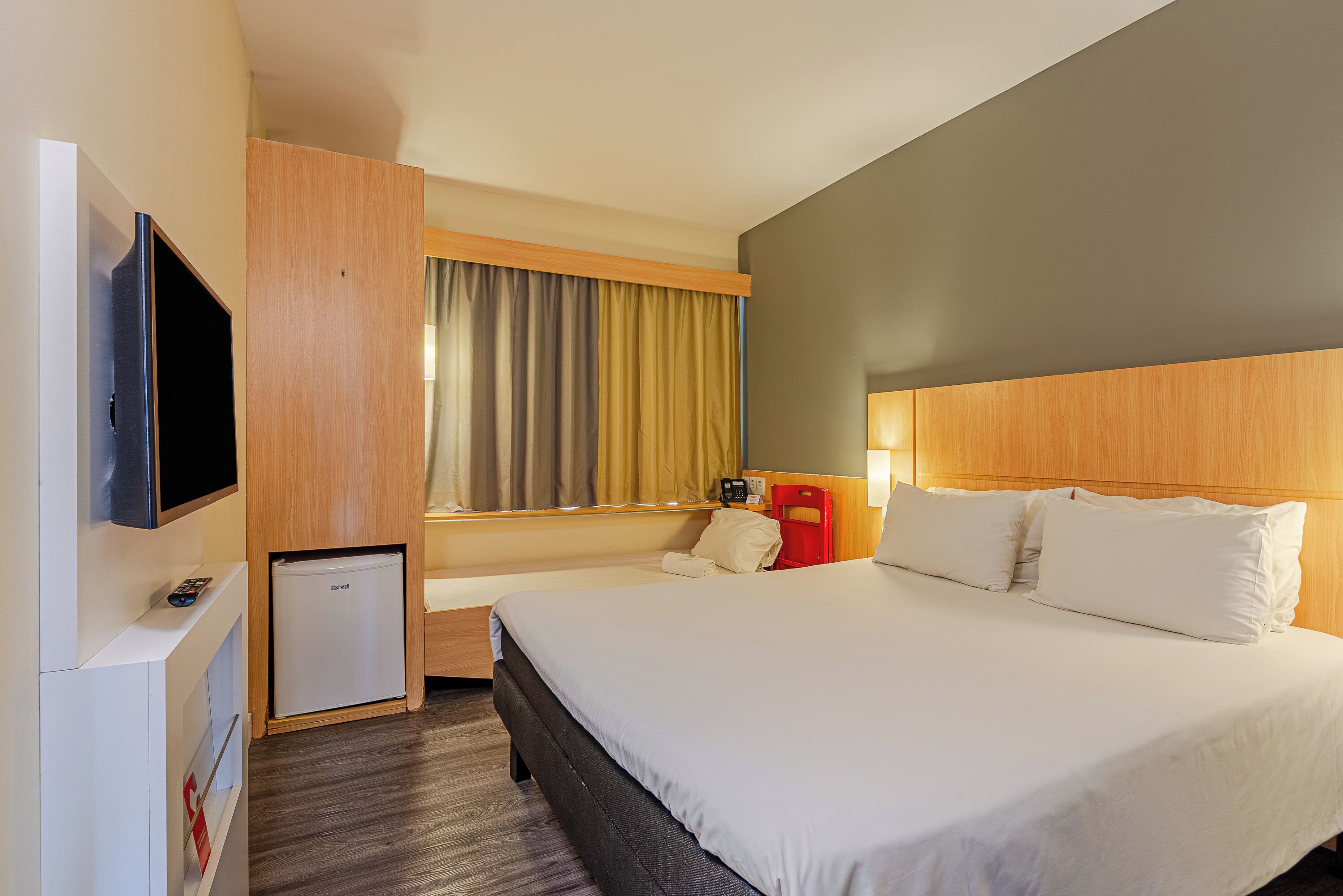 Quarto de hotel com cama, televisão, mini-geladeira e escrivaninha com luminária.