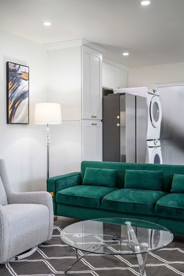 Uma sala de estar moderna com um sofá verde, uma poltrona cinza, uma mesa de centro de vidro, um abajur e uma máquina de lavar em segundo plano.