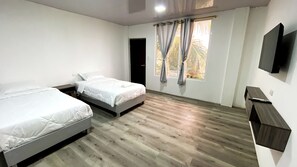 Quarto de hotel com duas camas de solteiro, piso de madeira, janela com cortinas e televisão de tela plana fixada na parede.