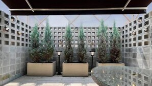 Uma área externa moderna com mesa de vidro, plantas em vasos e uma estrutura de parede geométrica.
