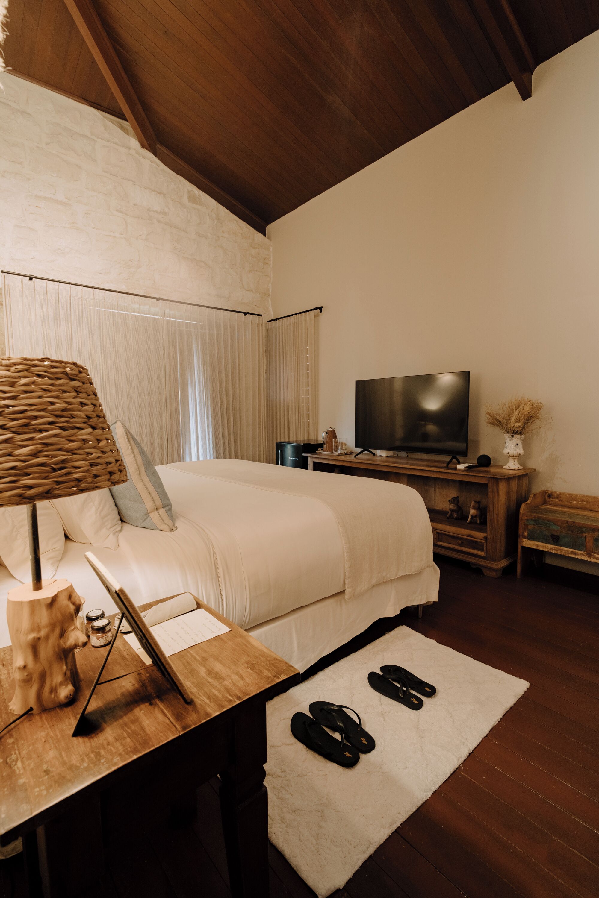 Quarto com cama grande, televisão, mesa de cabeceira de madeira e escrivaninha com abajur e um par de chinelos.