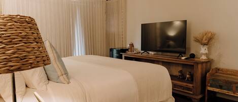 Quarto com cama grande, televisão, mesa de cabeceira de madeira e escrivaninha com abajur e um par de chinelos.
