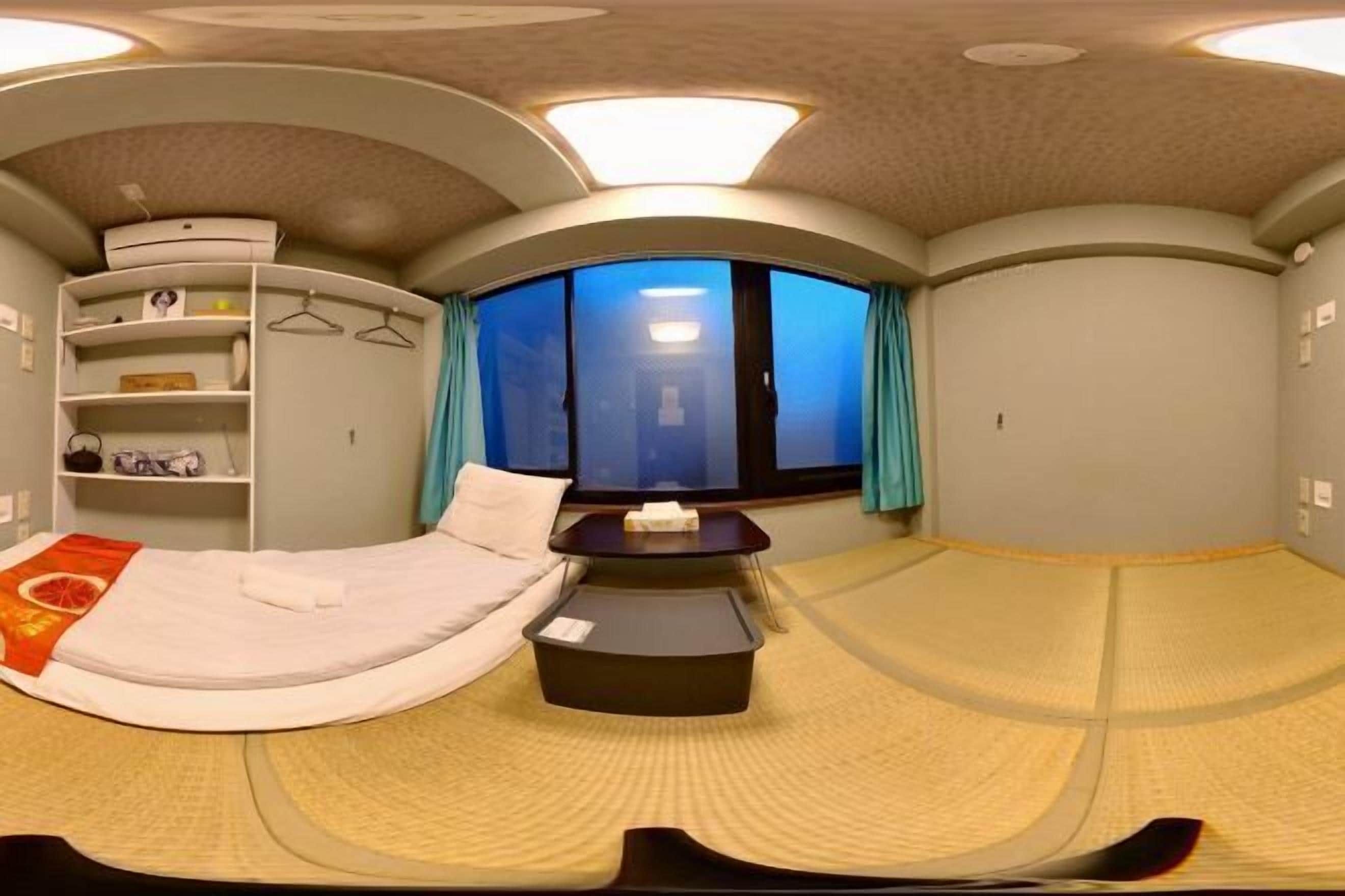 Quarto de hotel compacto com cama curva, uma pequena mesa, guarda-roupa e janela com cortinas.