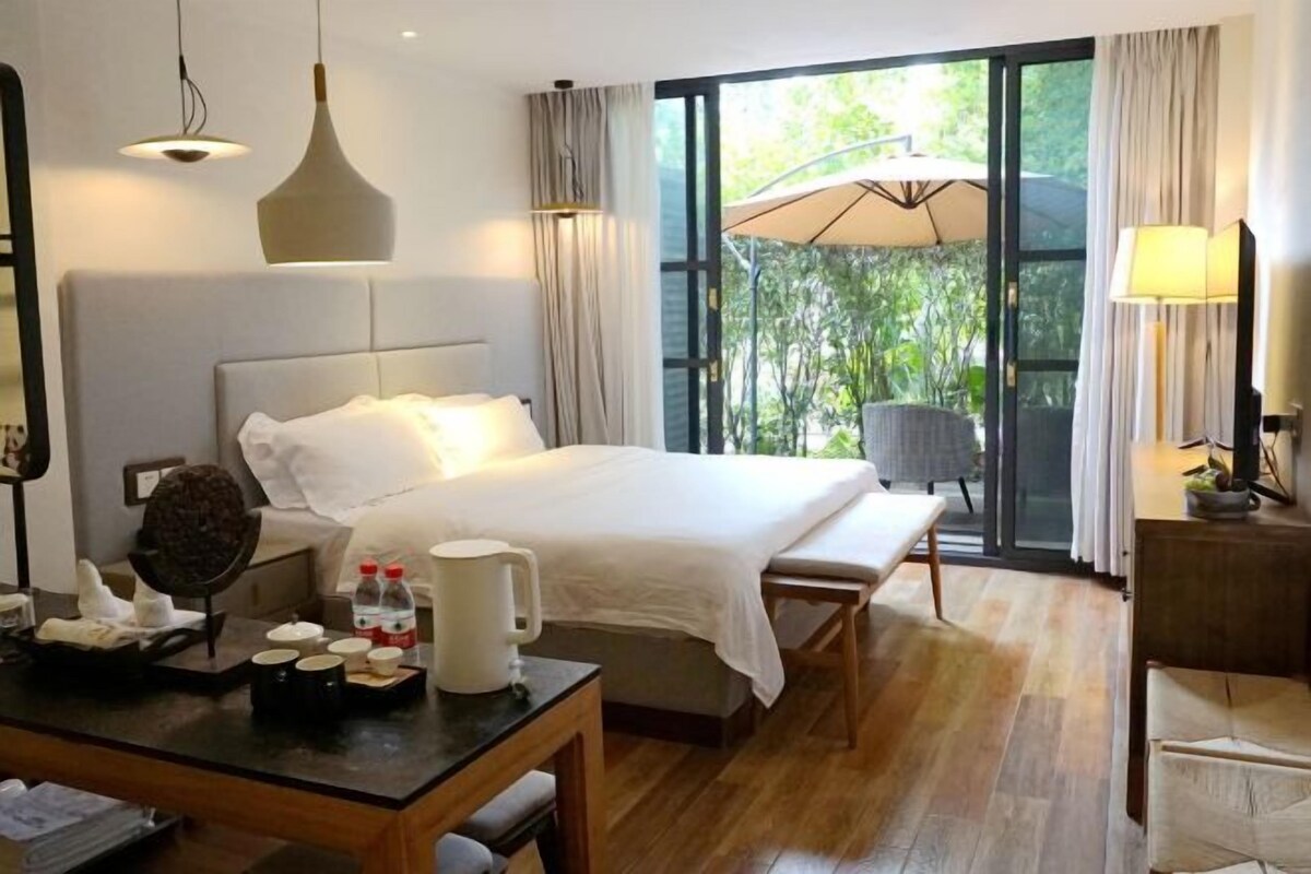 Quarto de hotel com cama, escrivaninha com luminária, cadeira e uma janela grande com cortinas.