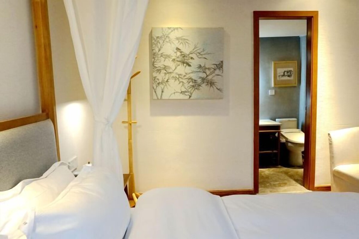 Quarto com cama, mesa de cabeceira, cadeira, quadro e banheiro visível por uma porta aberta.