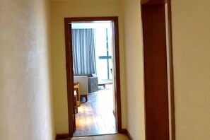 Corredor que dá para um quarto com uma cama, uma cadeira e uma janela com cortinas.