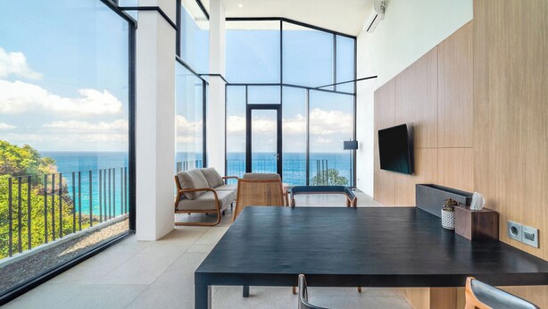 Quarto moderno com uma grande mesa preta, duas cadeiras, uma televisão e vista para o oceano.