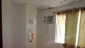 Quarto individual com cama de estrutura vermelha, colchão branco e toalha dobrada. Há uma janela com cortinas, um ar condicionado e um telefone fixado na parede.