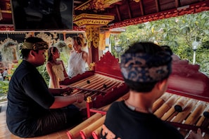 Interior de um templo balinês tradicional, com um músico sentado, uma mulher de vestido branco e um homem de camisa preta.