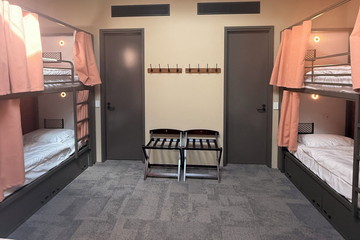 Quarto de dormitório com beliches, uma mesa central e um claraboia.