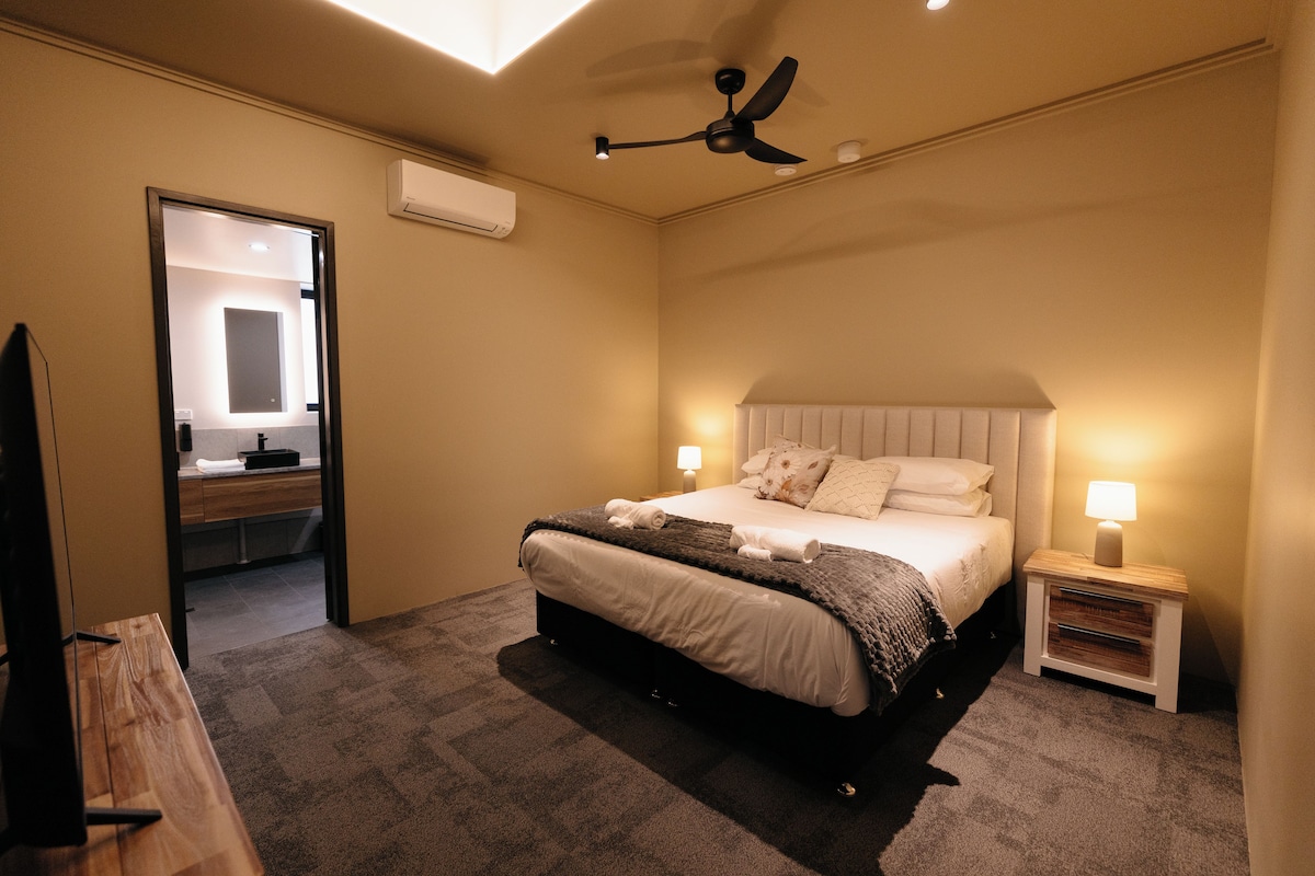 Quarto com cama, luminárias de cabeceira, ventilador de teto e espelho no banheiro próximo.