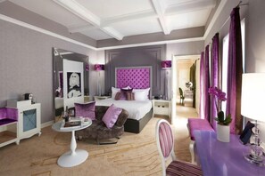 Quarto de hotel com uma cama grande, um espaço de estar com uma poltrona e uma mesinha, uma toucador com espelho e um banheiro visível através de uma porta aberta.