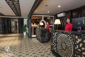 Um saguão de hotel com balcão de recepção, uma planta em vaso e um painel decorativo.