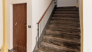 Uma escadaria com corrimão de madeira e balaústres pretos, que leva a um patamar com uma porta e uma janela com grades de metal.