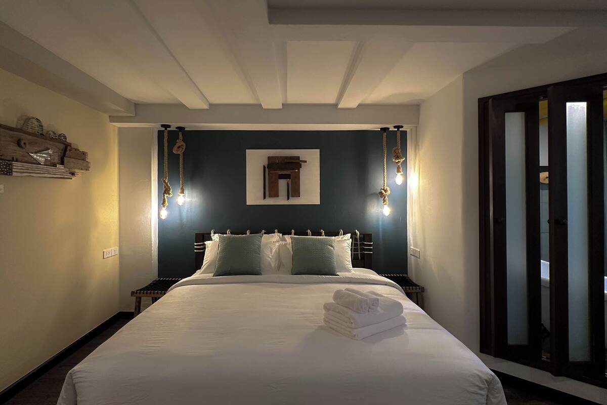 Quarto de hotel com cama, cabeceira com iluminação, prateleira de madeira e vista através de uma porta de vidro.