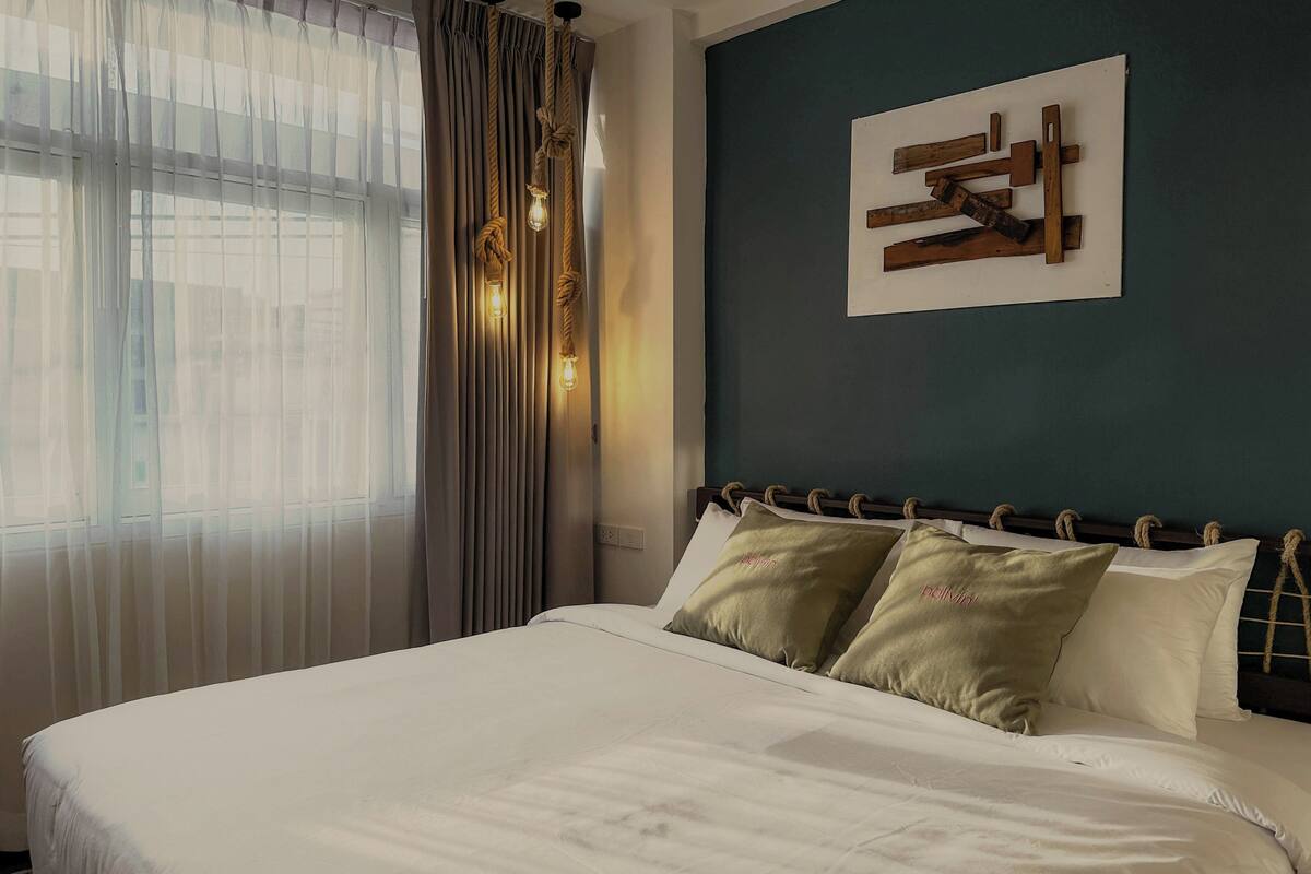 Cama bem arrumada com duas almofadas, parede escura com quadro e cortinas finas.