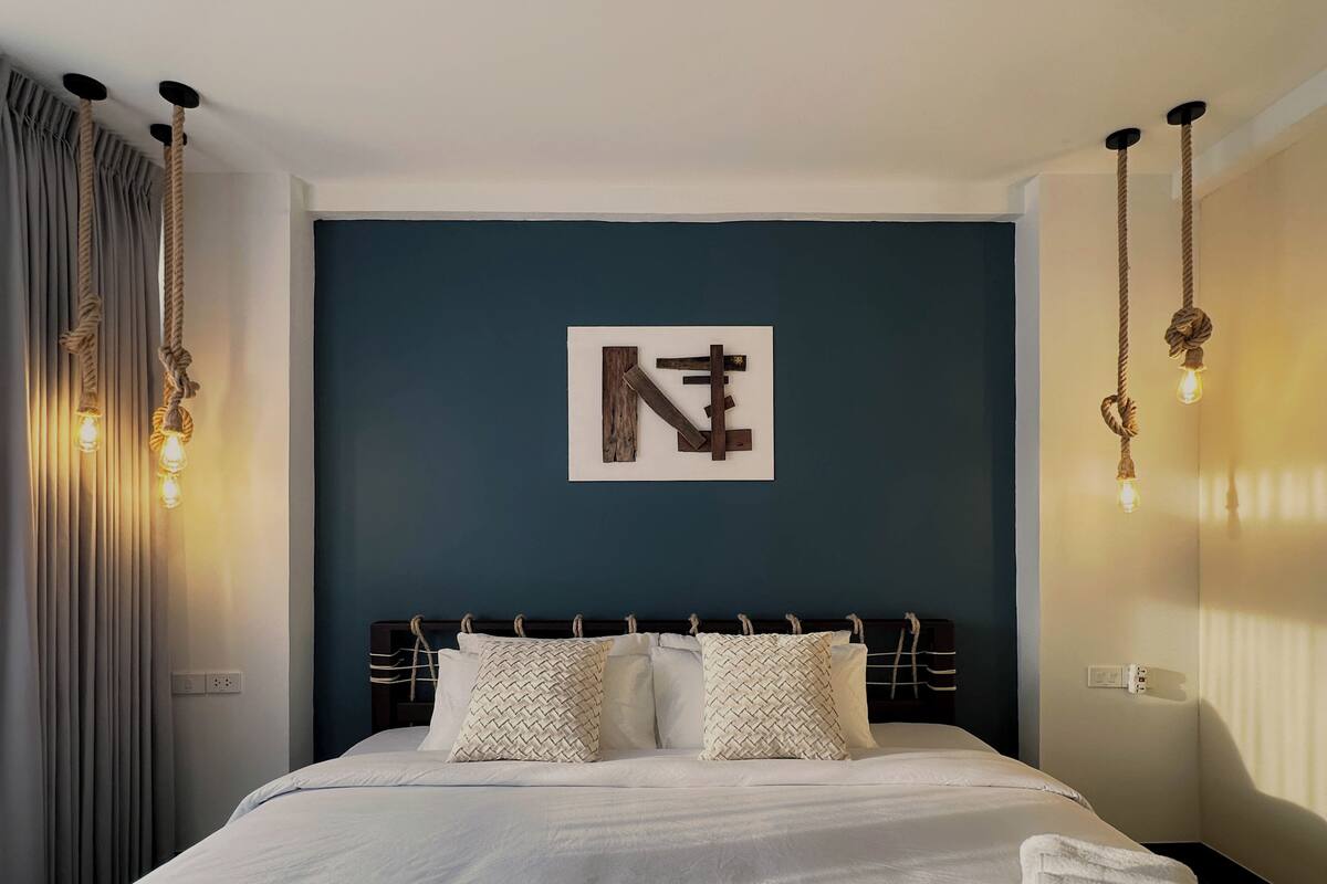 Uma cama bem arrumada com lençóis brancos e duas almofadas estampadas, uma parede azul escura de destaque e uma obra de arte emoldurada acima da cama.