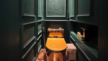 Um quarto estreito e pouco iluminado, com piso de madeira, um único vaso sanitário e uma pequena prateleira com objetos.