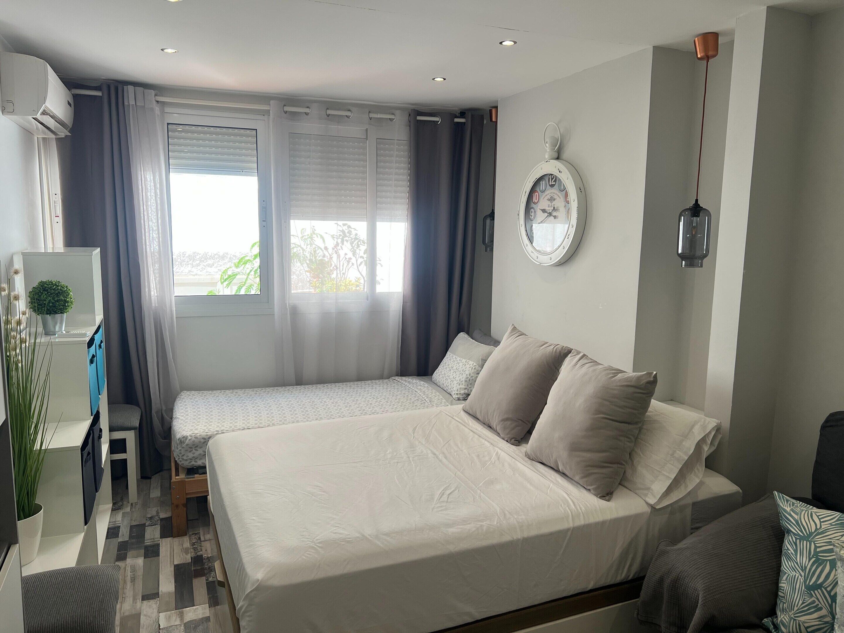 Quarto de hotel com duas camas, uma escrivaninha, um vaso de plantas e uma janela com cortinas.