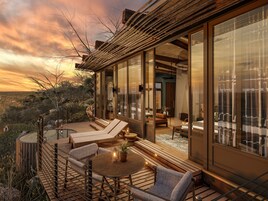 Uma área de estar moderna ao ar livre, com um deck de madeira, espreguiçadeiras e uma sala envidraçada com vista para a paisagem.